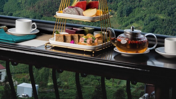 Uma mesa com bandeja escalonada de comida e chá, com paisagem montanhosa ao fundo.