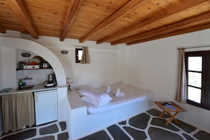 Quarto compacto com piso de pedra, pequena cozinha com pia e um refrigerador compacto, cama com roupa branca e janela com cortinas.