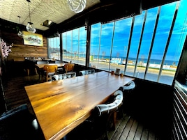 Um restaurante com uma grande mesa e cadeiras de madeira, um mural na parede e amplas janelas com vista para a praia e o oceano.