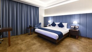 Quarto de hotel com uma cama grande, uma escrivaninha, duas cadeiras e uma mesa de cabeceira com luminárias.