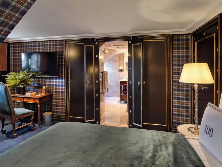 Quarto de hotel com uma cama, uma escrivaninha com cadeira, televisão e luminária.