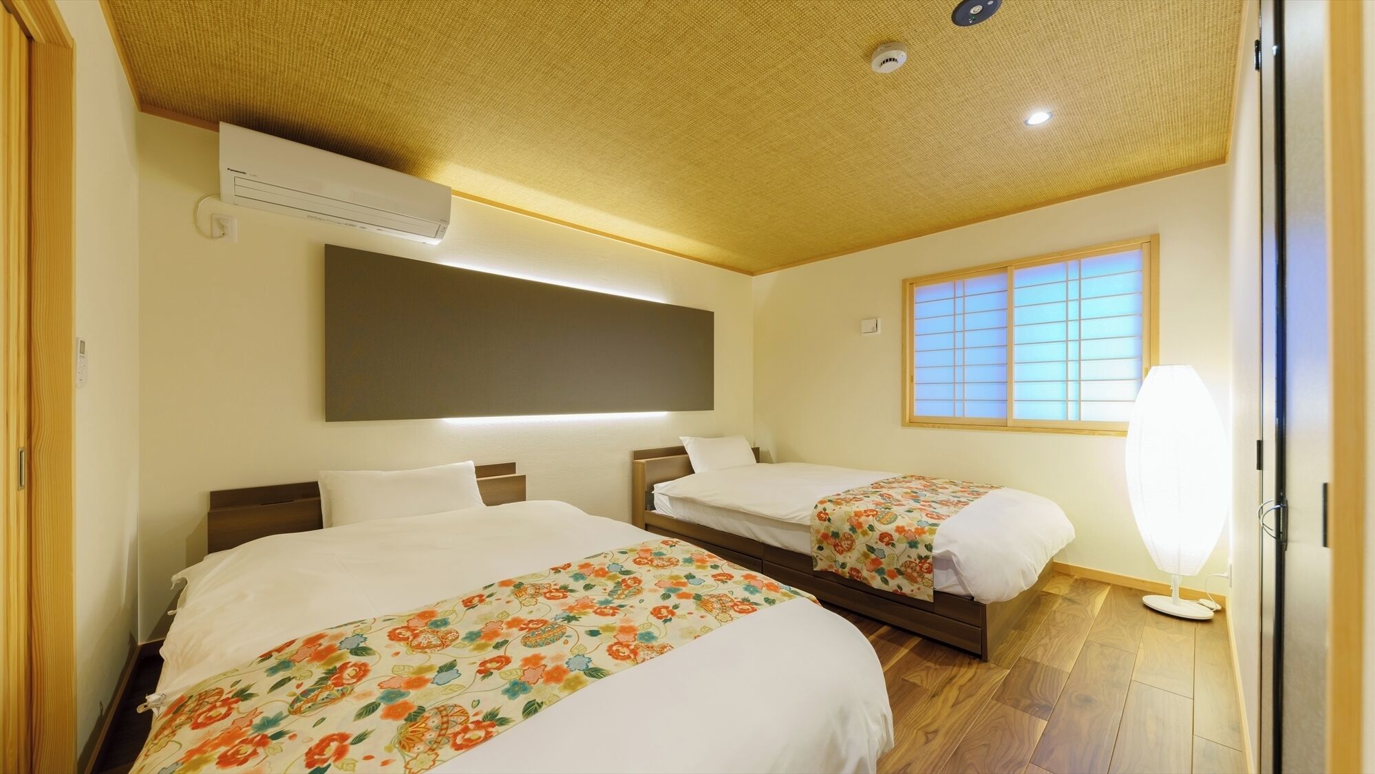Quarto de hotel com duas camas, uma TV grande fixada na parede, uma janela com persianas e um aparelho de ar condicionado.