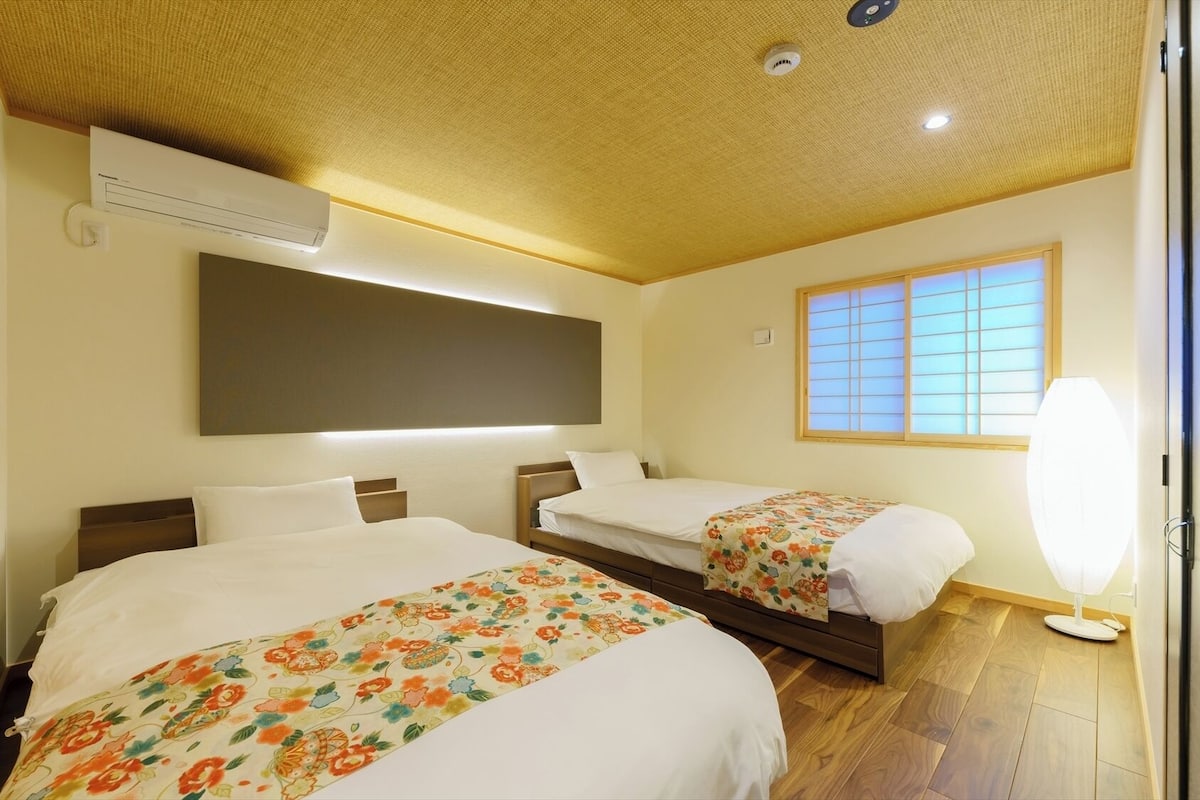 Quarto de hotel com duas camas, uma TV grande fixada na parede, uma janela com persianas e um aparelho de ar condicionado.