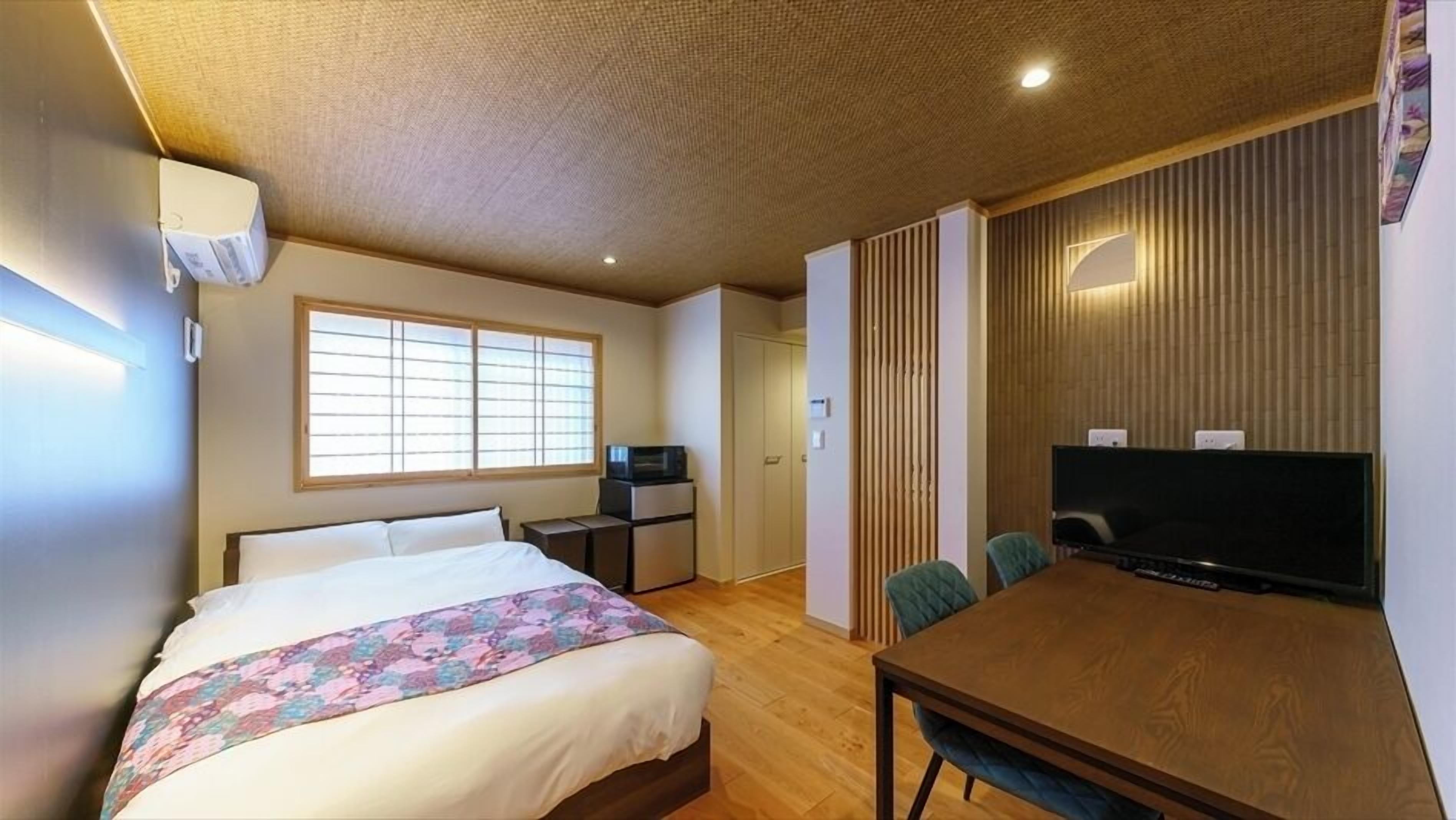 Quarto de hotel com cama, escrivaninha com computador, televisão e mesa de jantar com cadeiras.
