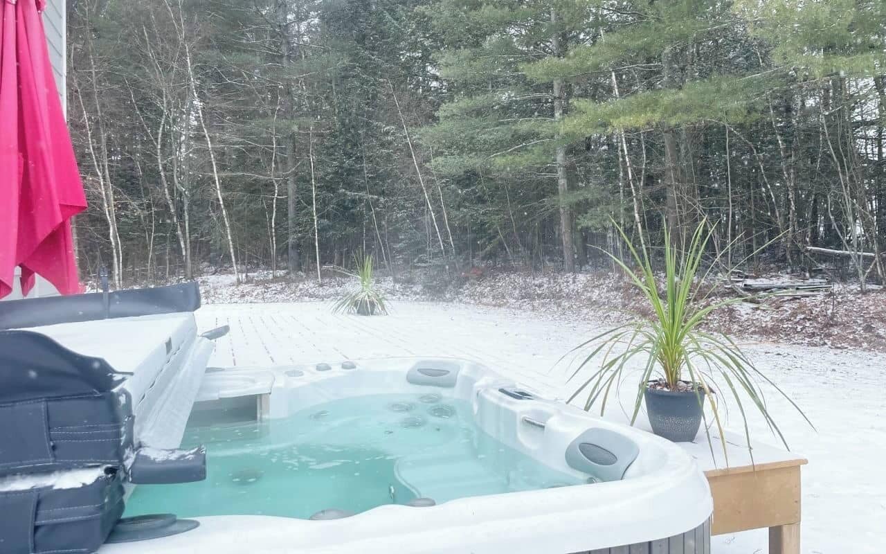 Uma banheira de hidromassagem numa área externa nevada com deck de madeira, rodeada por árvores.