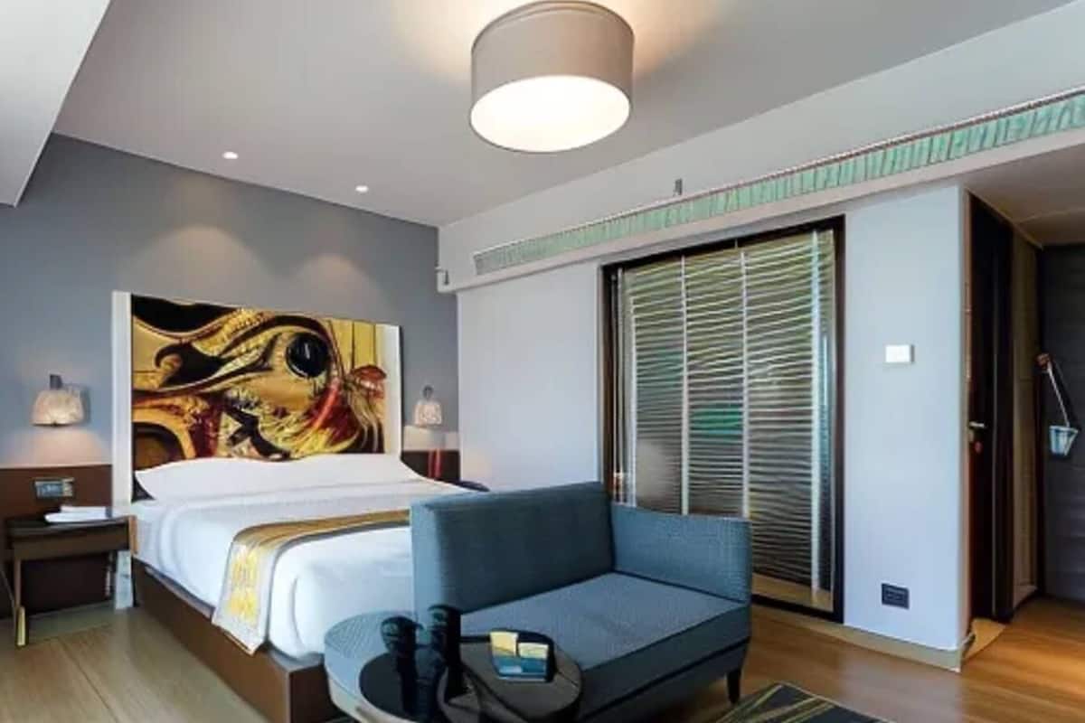 Quarto de hotel moderno com uma cama grande, um sofá azul, uma obra de arte na parede, uma mesa de cabeceira com abajur e um guarda-roupa com portas de correr.