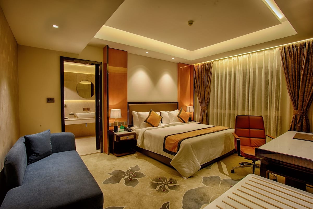Quarto de hotel com uma cama grande, uma mesa, uma cadeira, um sofá, um espelho e um banheiro visível por uma porta aberta.