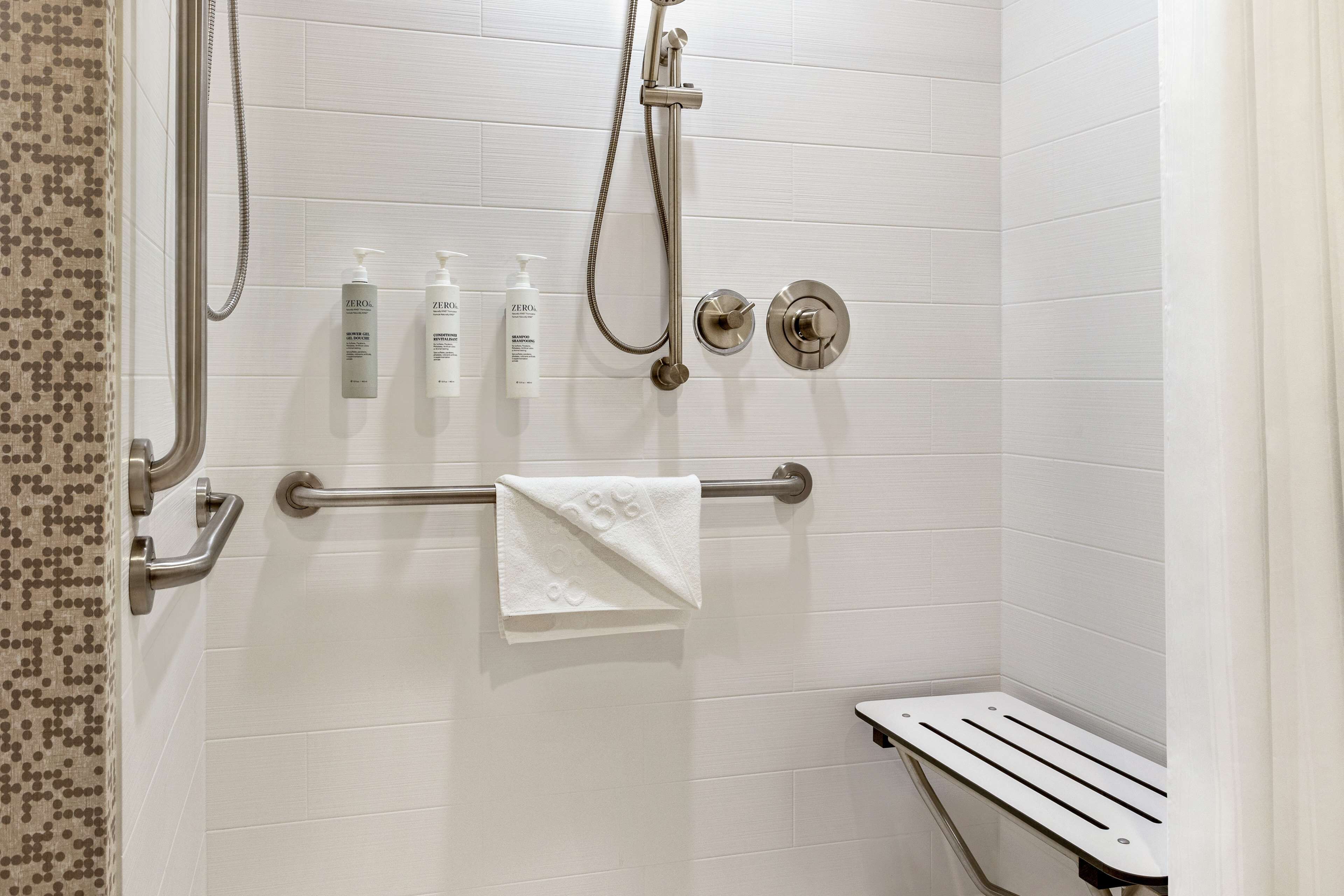 Uma área de chuveiro com paredes em azulejos brancos, um chuveiro manual, um porta-toalhas com uma toalha branca e duas garrafas de shampoo.