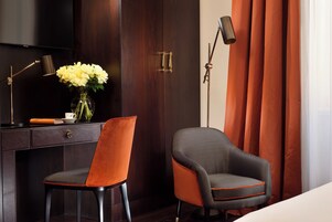 Quarto de hotel com escrivaninha, duas cadeiras, um vaso com flores, luminária e cortinas.