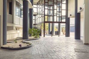 Um espaço amplo e interno com grandes janelas, colunas e piso ladrilhado.