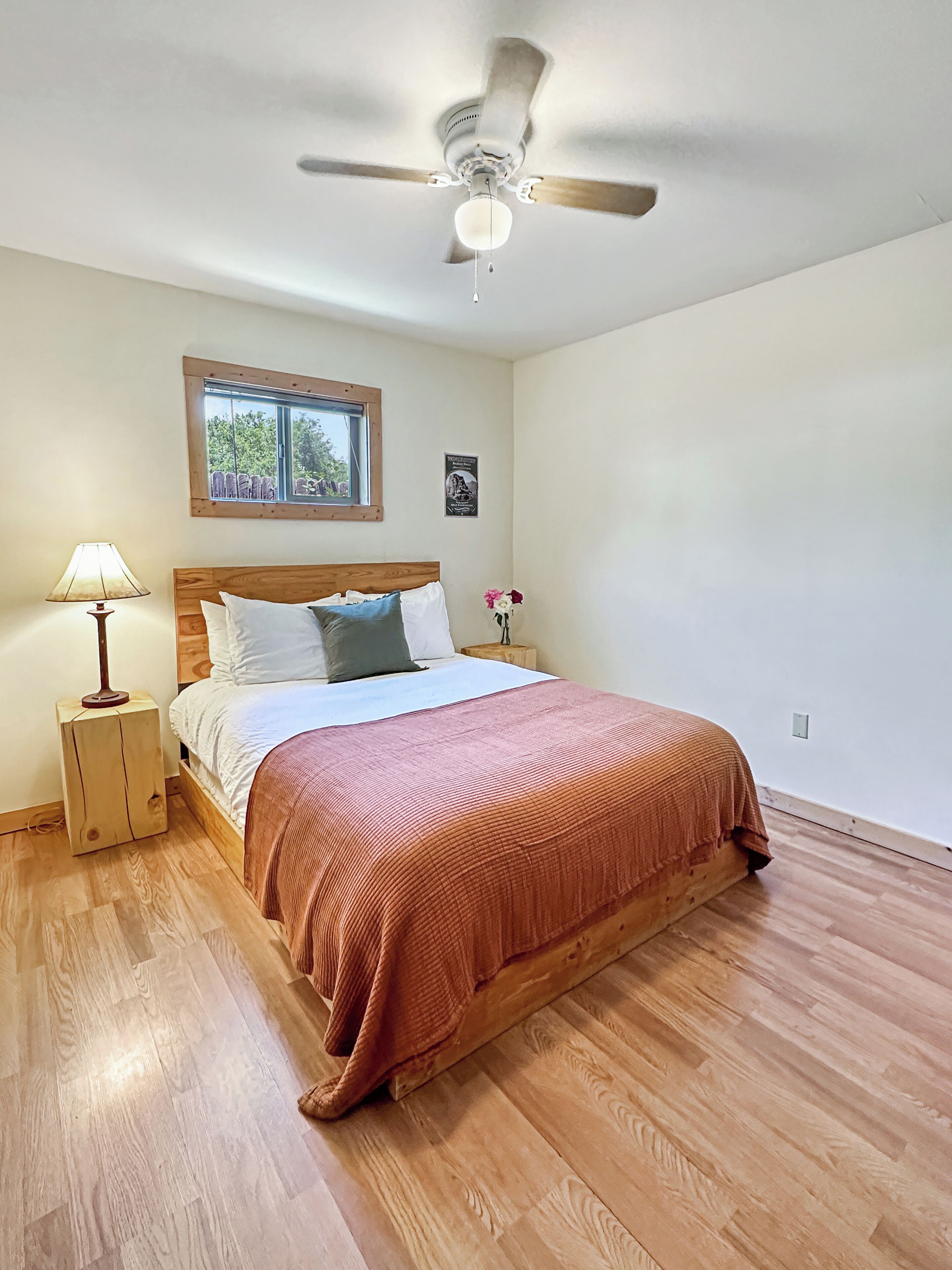 Um quarto com cama, ventoinha no teto, janela, mesa de cabeceira e abajur.