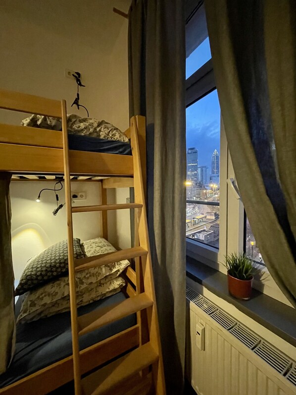 Uma cama beliche com escada, um vaso de planta no parapeito da janela e vista para a cidade através da janela.