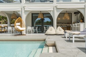 Área na piscina com uma cadeira suspensa, sofás brancos e um bar com vista para a piscina.