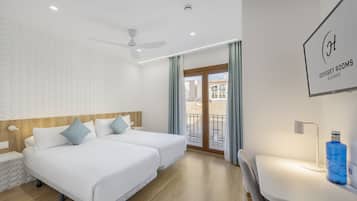 Quarto de hotel com duas camas, uma escrivaninha, uma cadeira, ventoinha no teto e vista para a cidade através de uma varanda.