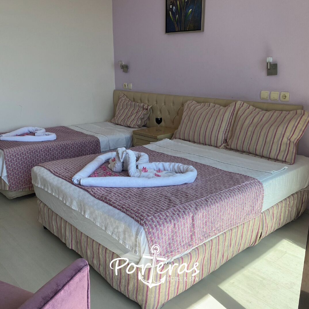Quarto de hotel com duas camas, cada uma com colcha listrada em rosa e branco, uma cama grande com padrão semelhante e uma mesinha com uma taça de vinho.