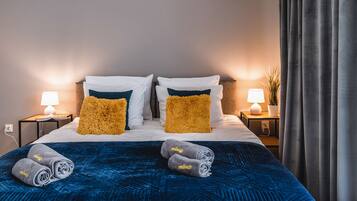 Cama bem arrumada com manta azul, dois travesseiros e toalhas enroladas sobre um cobre-leito azul escuro. Há duas mesas de cabeceira com luminárias e um vaso com planta.