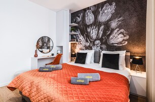 Quarto de hotel moderno com uma cama grande, um mural na parede, uma mesa de cabeceira com luminária e uma pequena área de estar com uma poltrona e mesa lateral.