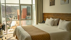 Quarto de hotel com uma cama grande, uma mesa e uma cadeira. Há uma janela com vista para a cidade e montanhas.