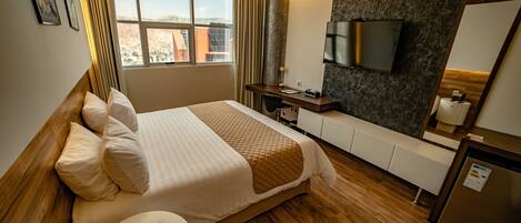 Quarto de hotel com cama, mesa de cabeceira, escrivaninha, televisão e vista para a cidade.