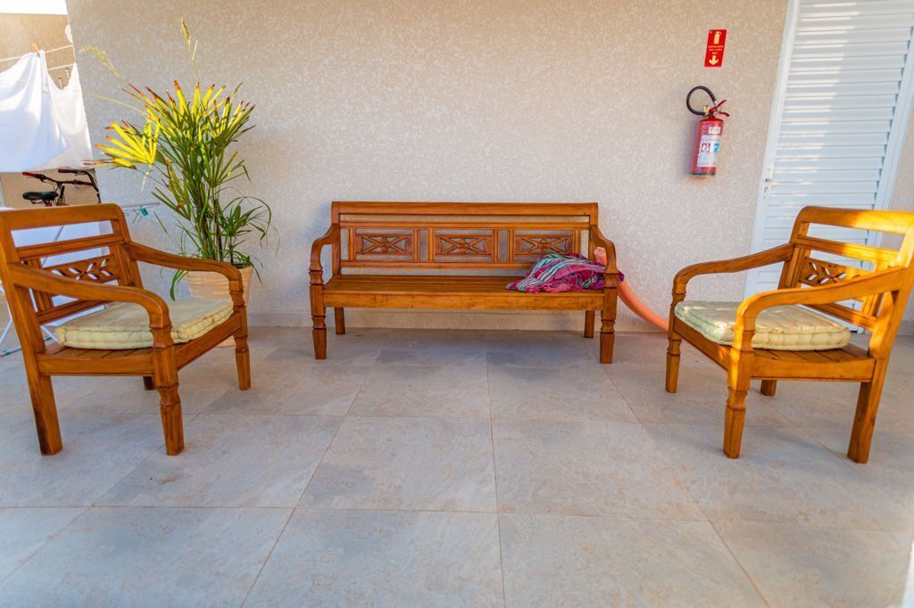 Um banco de madeira com almofada, ladeado por duas cadeiras de madeira também com almofadas, em um quarto com piso de azulejos e um extintor de incêndio fixado na parede.