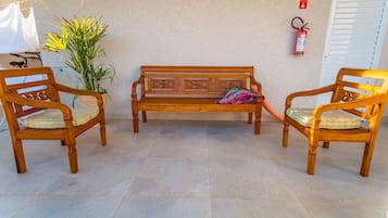 Um banco de madeira com almofada, ladeado por duas cadeiras de madeira também com almofadas, em um quarto com piso de azulejos e um extintor de incêndio fixado na parede.