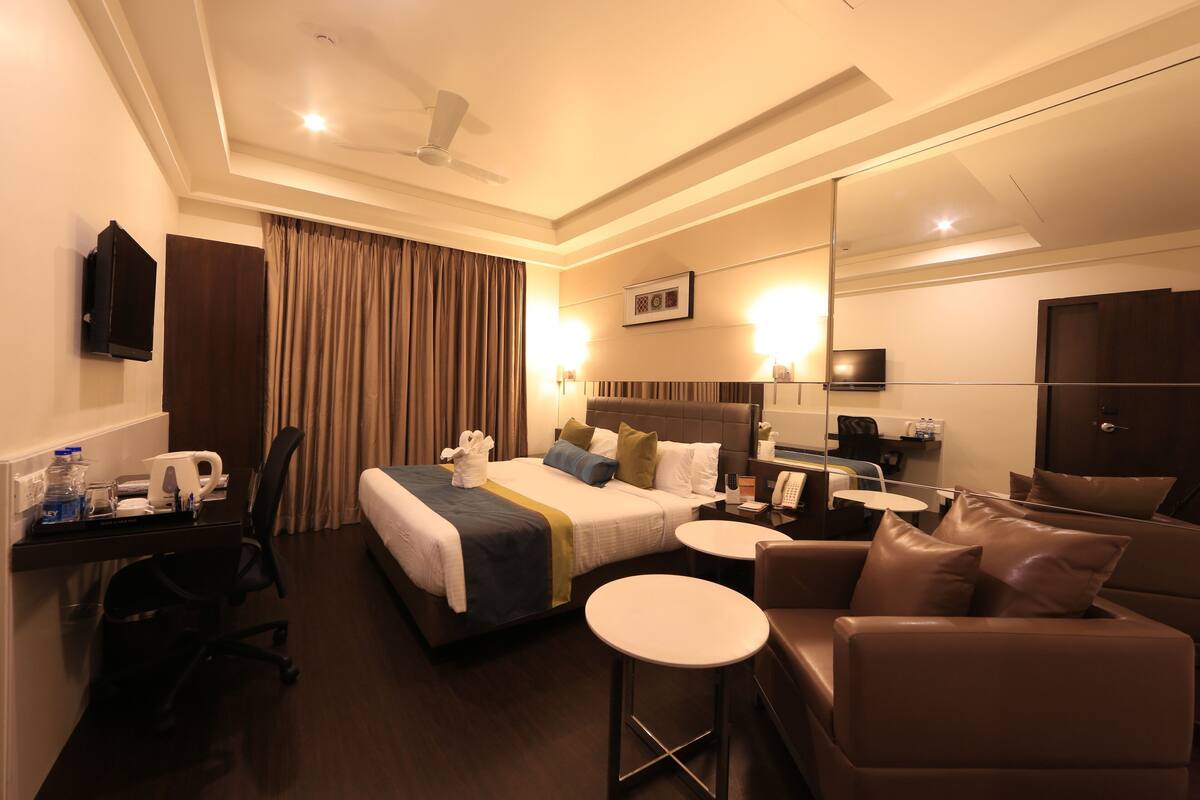 Quarto de hotel com uma cama, duas mesas redondas, um sofá, uma escrivaninha com cadeira, uma televisão e um ventilador de teto.