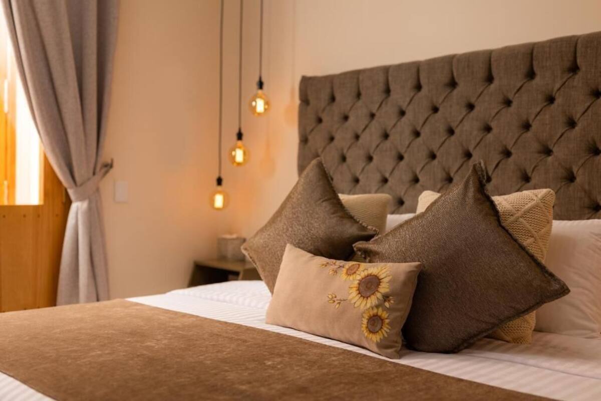 Cama bem arrumada com cabeceira estofada, vários travesseiros e manta marrom.