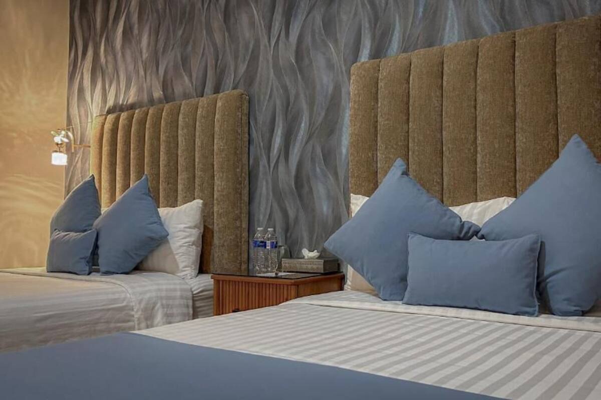 Quarto de hotel com duas camas, uma cabeceira grande e uma mesinha de cabeceira com uma garrafa de água.