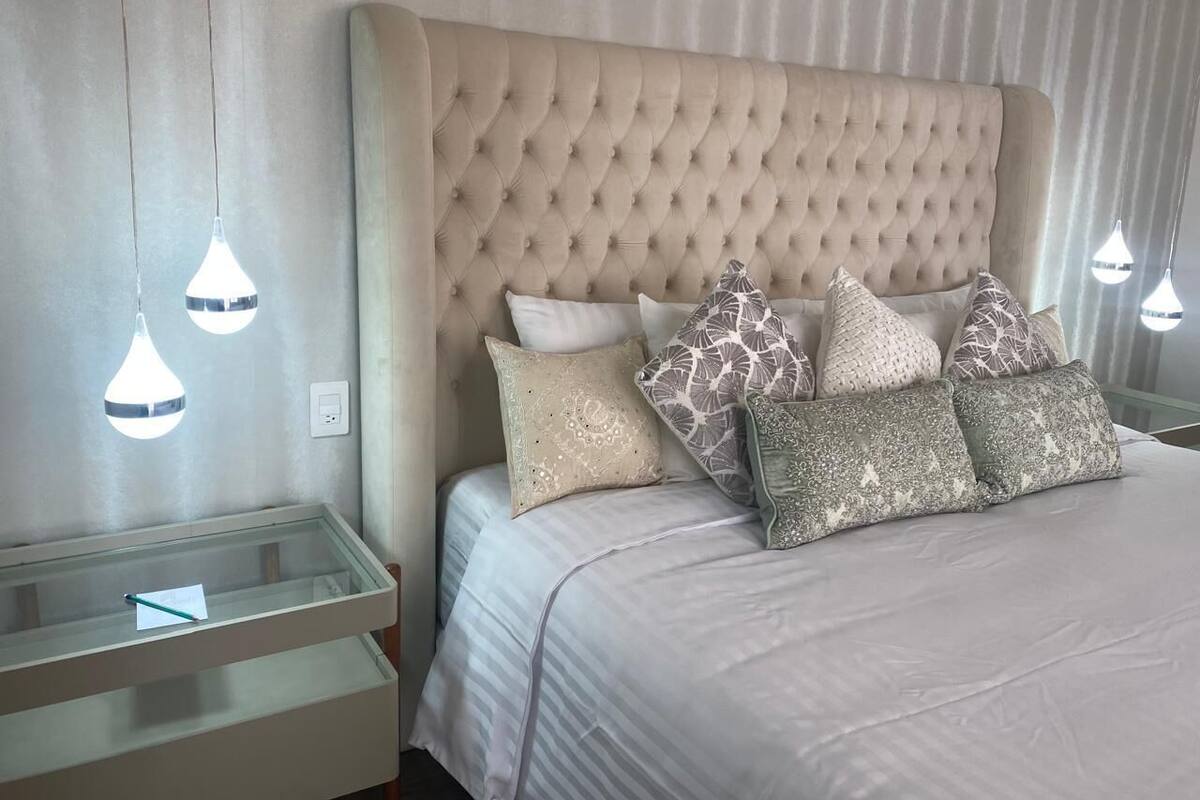 Quarto com cabeceira estofada, mesa de cabeceira de vidro e cama com almofadas estampadas.