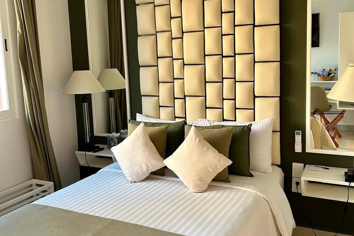 Quarto de hotel com uma cama grande, cabeceira com padrão geométrico, mesa de cabeceira com abajur e um espelho na parede.