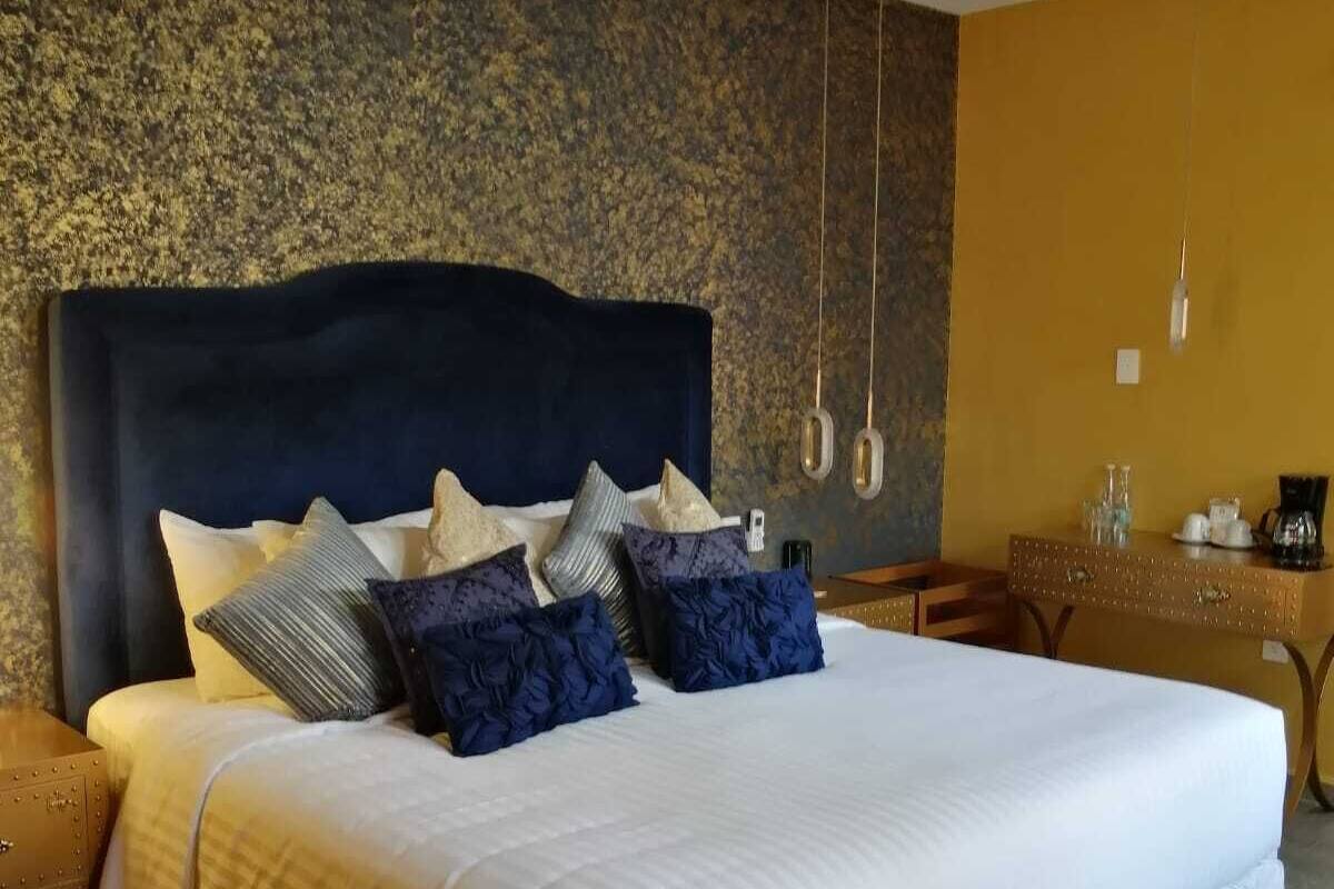 Uma cama grande com cabeceira escura, várias almofadas e uma mesa de cabeceira com abajur.