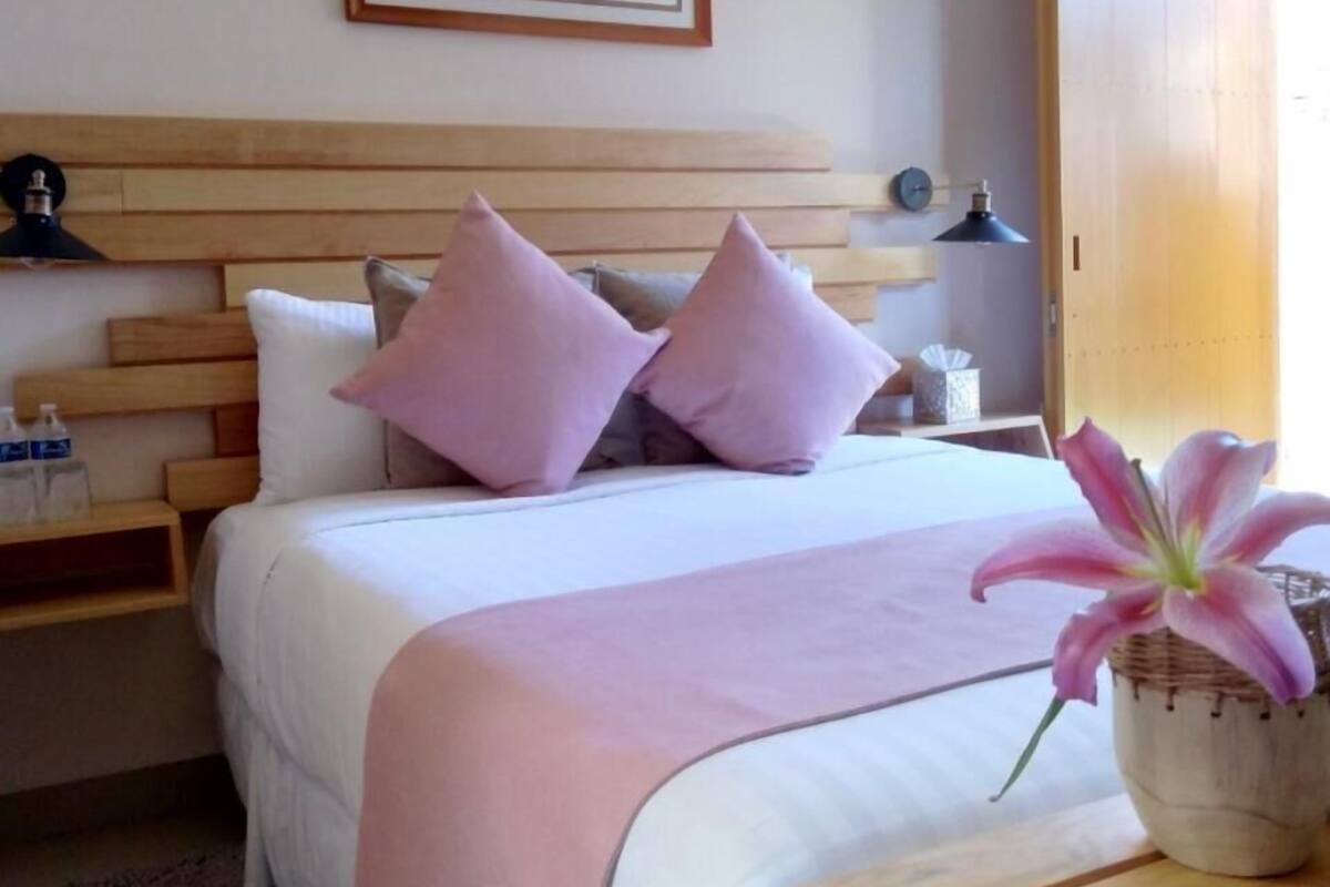 Quarto bem arrumado com cabeceira de madeira, cama com roupa de cama branca e almofadas cor-de-rosa, mesa de cabeceira com abajur, quadro na parede e vaso com uma flor cor-de-rosa em um criado-mudo de madeira.