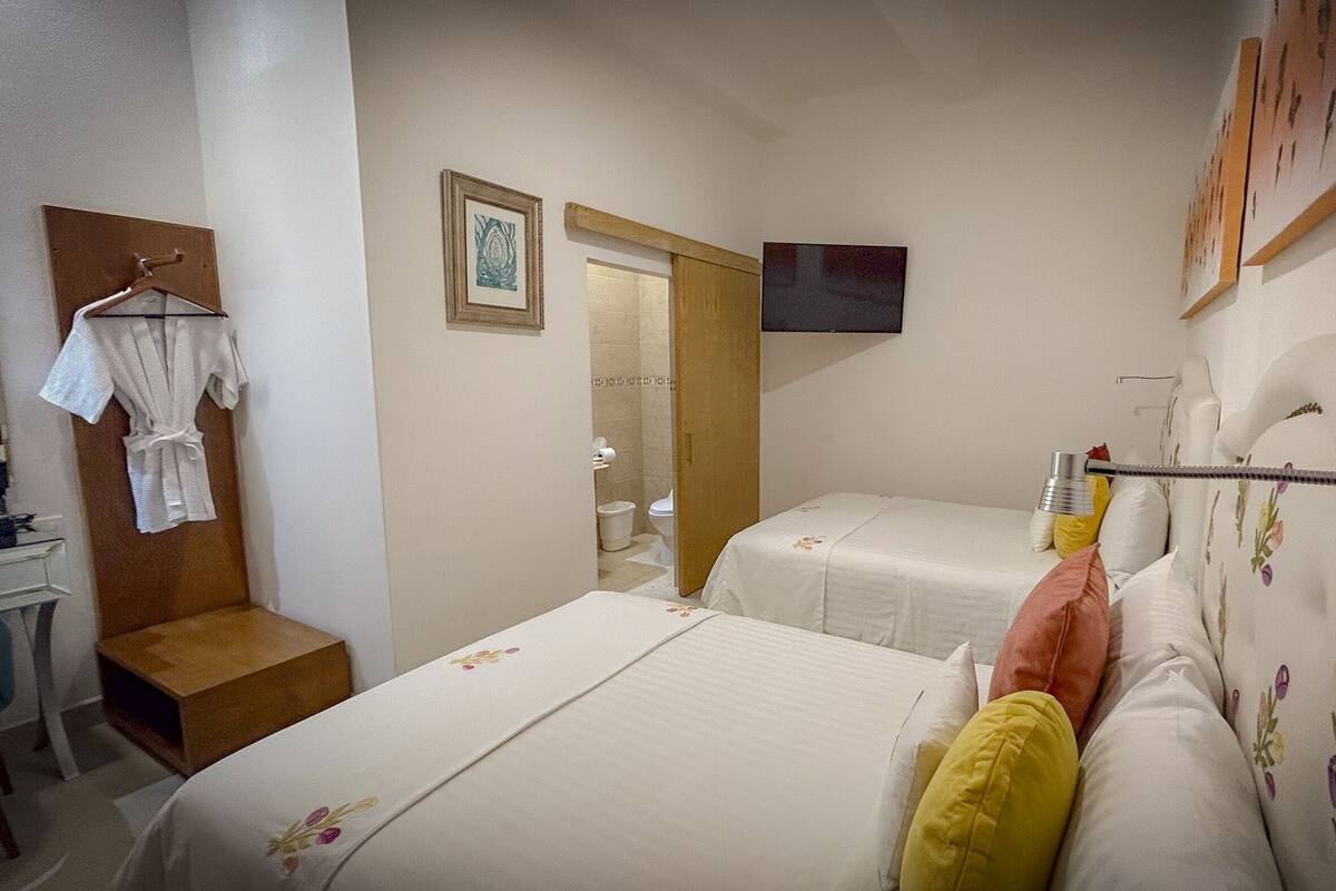 Quarto de hotel com duas camas, um banco de madeira, suporte para roupões e banheiro visível por uma porta aberta.