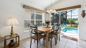 Uma área de refeições com mesa e cadeiras de madeira, uma luminária e vista para uma piscina através de grandes janelas.