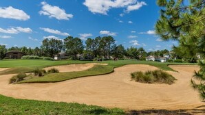 Um campo de golfe com armadilhas de areia, céu claro e edifícios residenciais ao fundo.