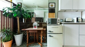 Uma cozinha compacta com geladeira branca, micro-ondas e torradeira. Há uma mesa de madeira com um cata-vento branco sobre ela, e uma planta em vaso à esquerda.