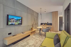 Uma sala de estar moderna com uma TV de tela plana na parede, um banco de madeira, um sofá verde e uma área de jantar com mesa redonda e cadeiras.