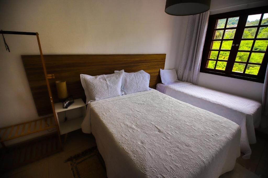 Quarto com uma cama grande, uma cama menor, cabeceira de madeira, mesa de cabeceira, abajur e janela com vista para vegetação.