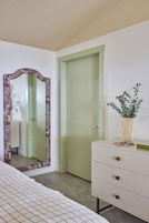 Quarto com cama, penteadeira, espelho com moldura floral, porta verde e planta em vaso.