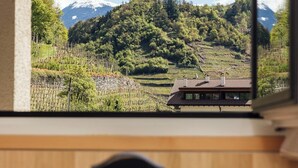 Visão pela janela de um quarto com escrivaninha e cadeira, com vista para uma paisagem montanhosa com casa e vinhedo.