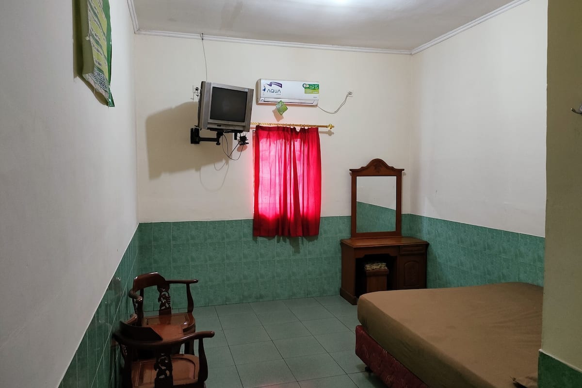 Quarto pequeno e simples com uma cama, uma cadeira de madeira, uma mesinha, um espelho, uma televisão e uma cortina vermelha.