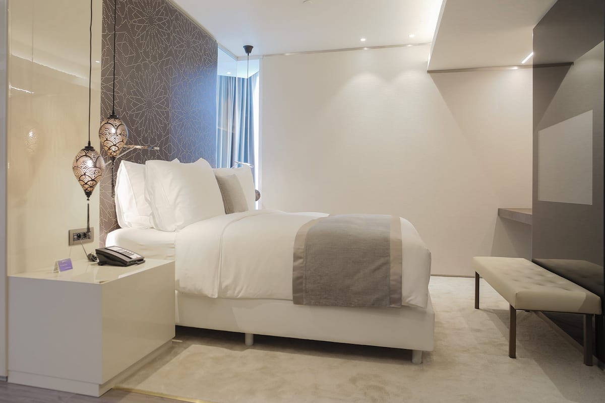 Quarto moderno com cama, mesa de cabeceira, luminária e banqueta.