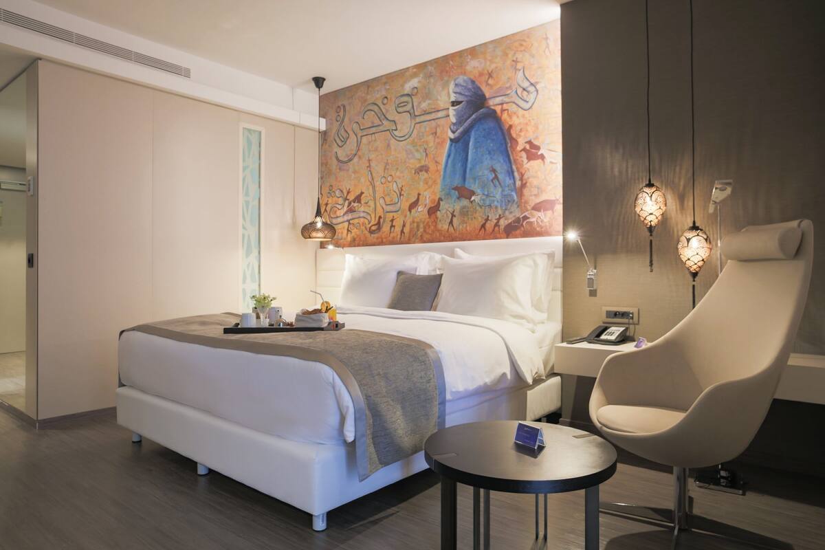 Quarto de hotel moderno com uma cama, uma cadeira, uma mesinha e uma parede com caligrafia árabe.