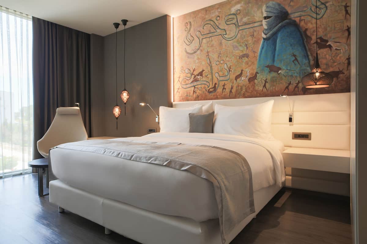 Quarto moderno com uma cama grande, mesa de cabeceira, uma cadeira e um mural na parede.