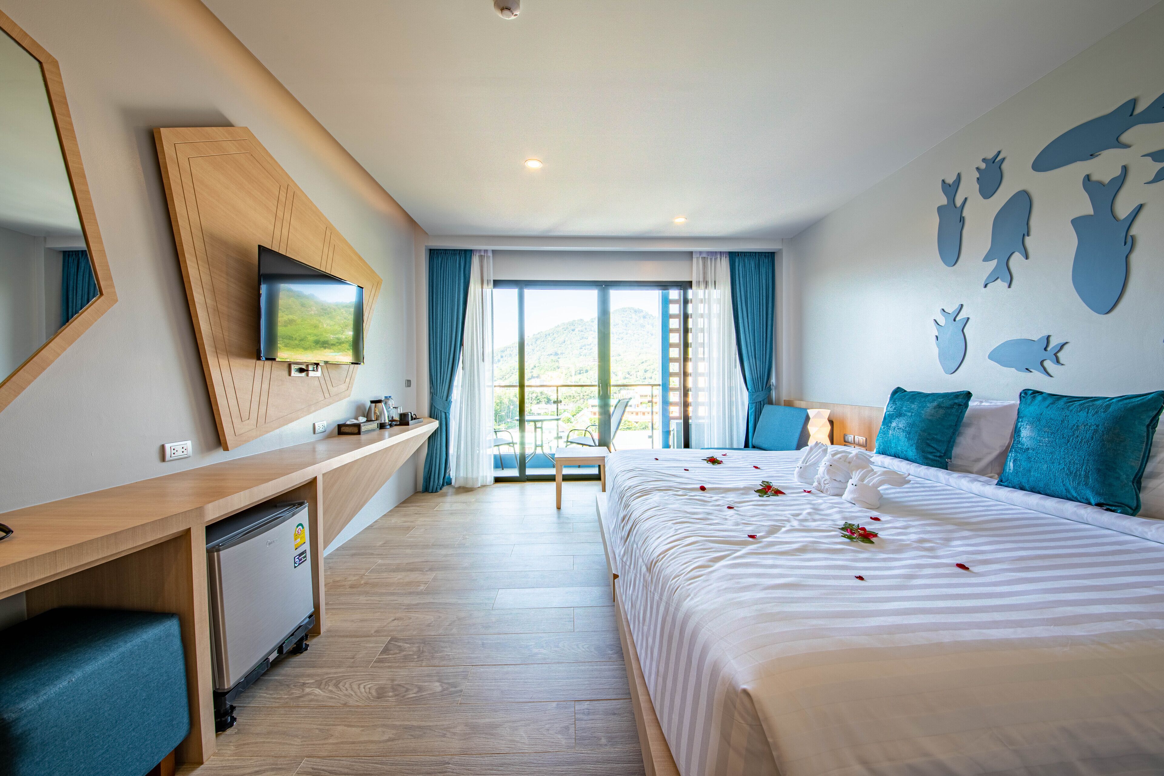 Quarto de hotel com cama, escrivaninha, mini-geladeira, televisão e uma grande janela com cortinas.