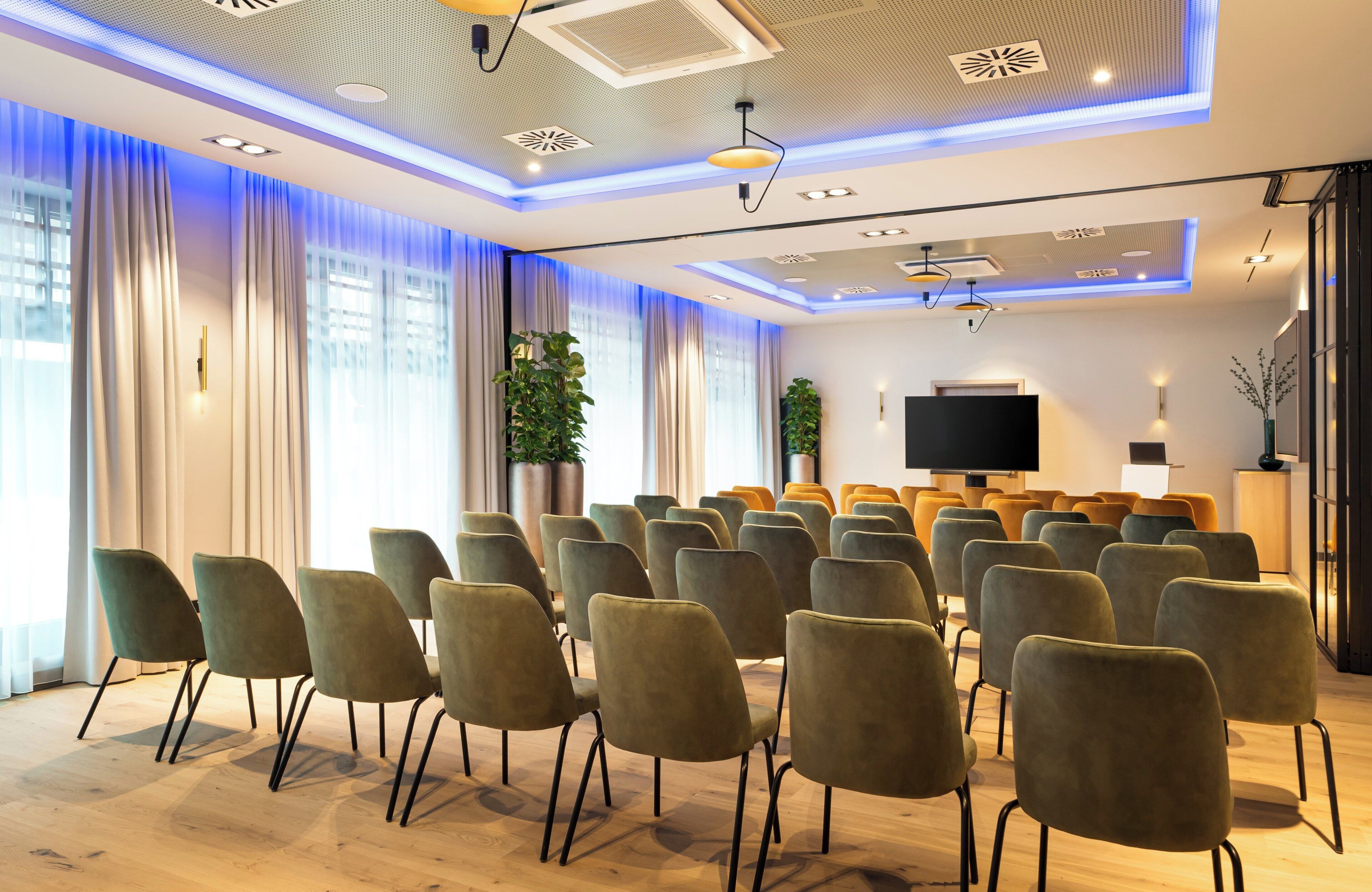 Uma sala de conferências com fileiras de cadeiras, uma tela grande e uma planta.