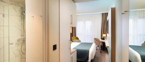 Quarto de hotel com chuveiro, escrivaninha com cadeira, cama e janela com cortinas.
