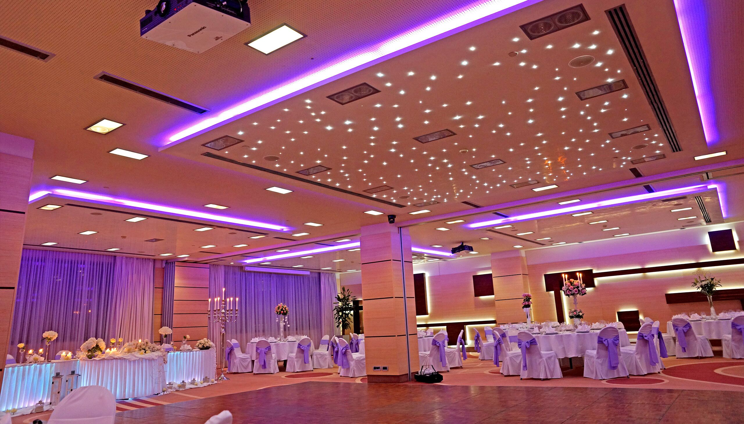 Salão de banquetes com mesas montadas para um evento formal, com luminárias no teto e área de palco.