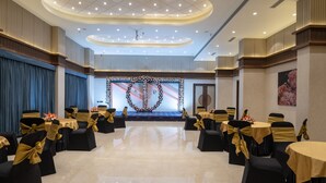 Uma ampla sala de banquetes com palco, mesas redondas e cadeiras pretas com fitas douradas.