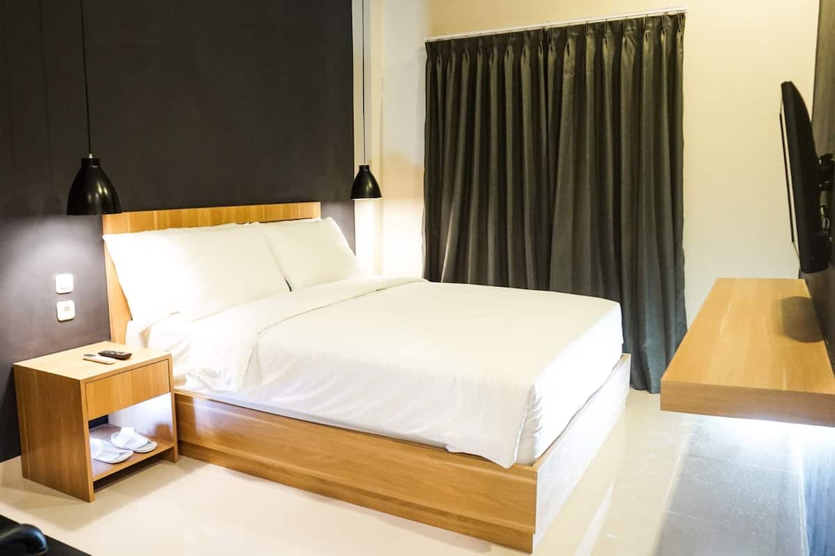Quarto de hotel moderno com estrutura de cama de madeira, roupa de cama branca, mesa de cabeceira com abajur e TV sobre suporte de madeira.